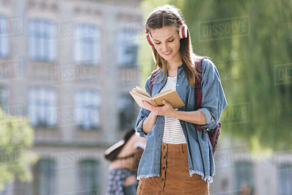 Happy attractive female student with headphones reading book near ...