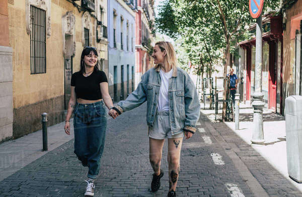 Cheerful Lesbians Holding Hands While Walking On Blurred Background Of