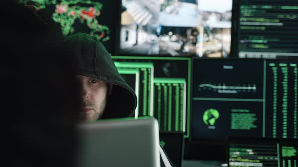 Hooded Computer hacker coding on computer while sitting at office full of monitor screens ...