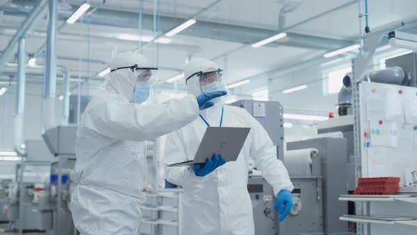 Two Scientists Standing in a Heavy Industry Factory in Sterile Coveralls and Face Masks, Using ...