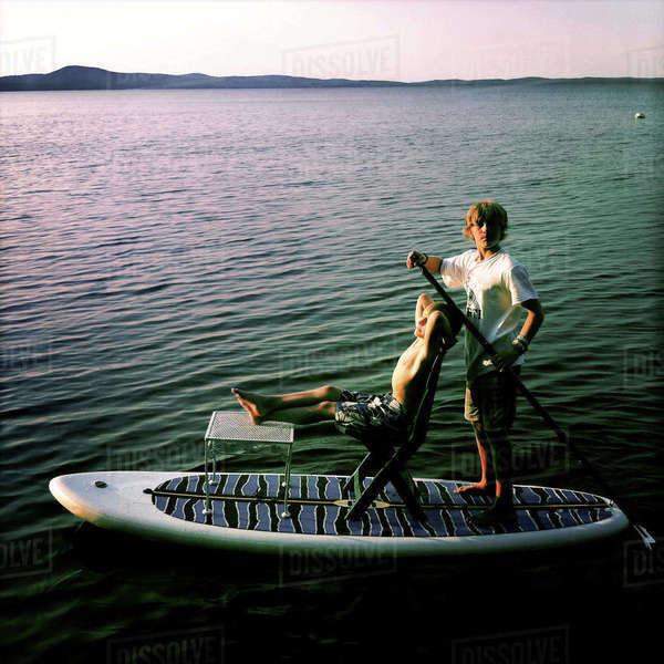 An eleven year old boy paddles while his eight year old brother relaxes on their standup paddle ...
