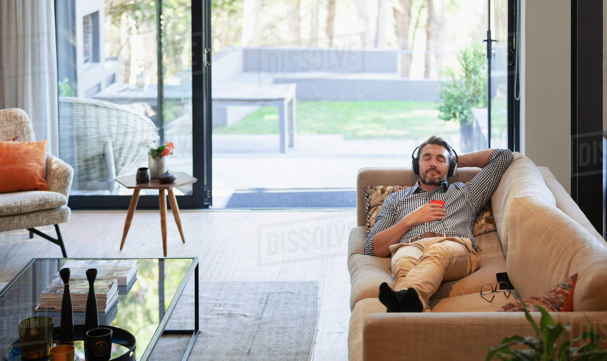 Man relaxing on living room sofa, listening to music with headphones ...
