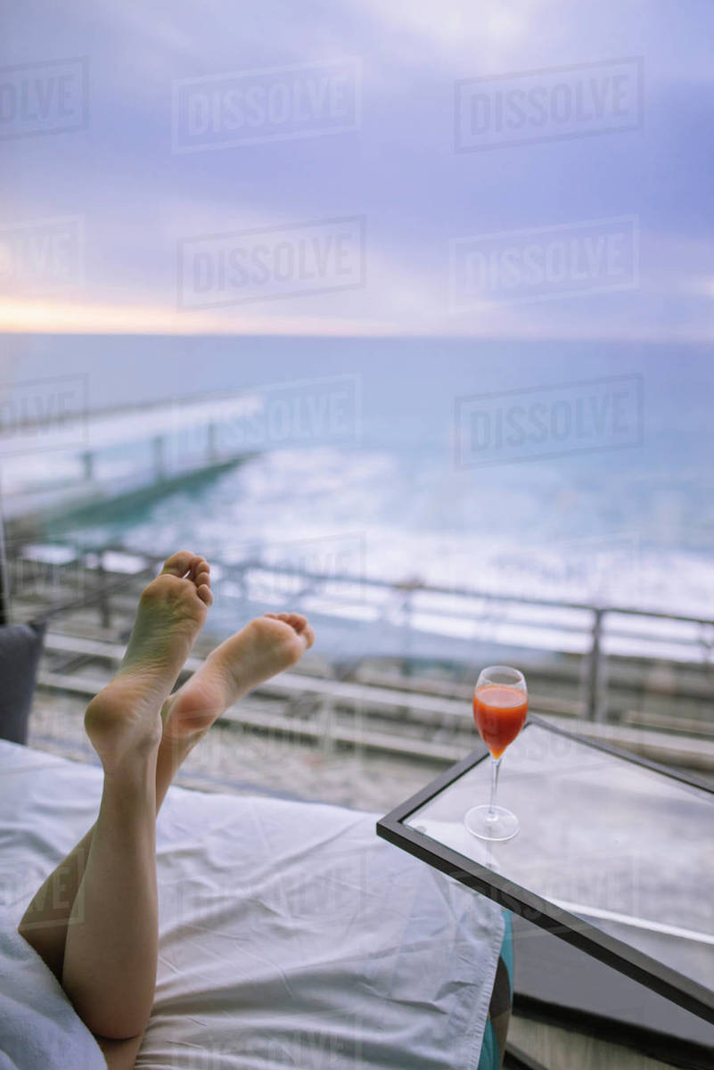 Woman's feet and cocktail in hotel room with view on sea at sunrise ...