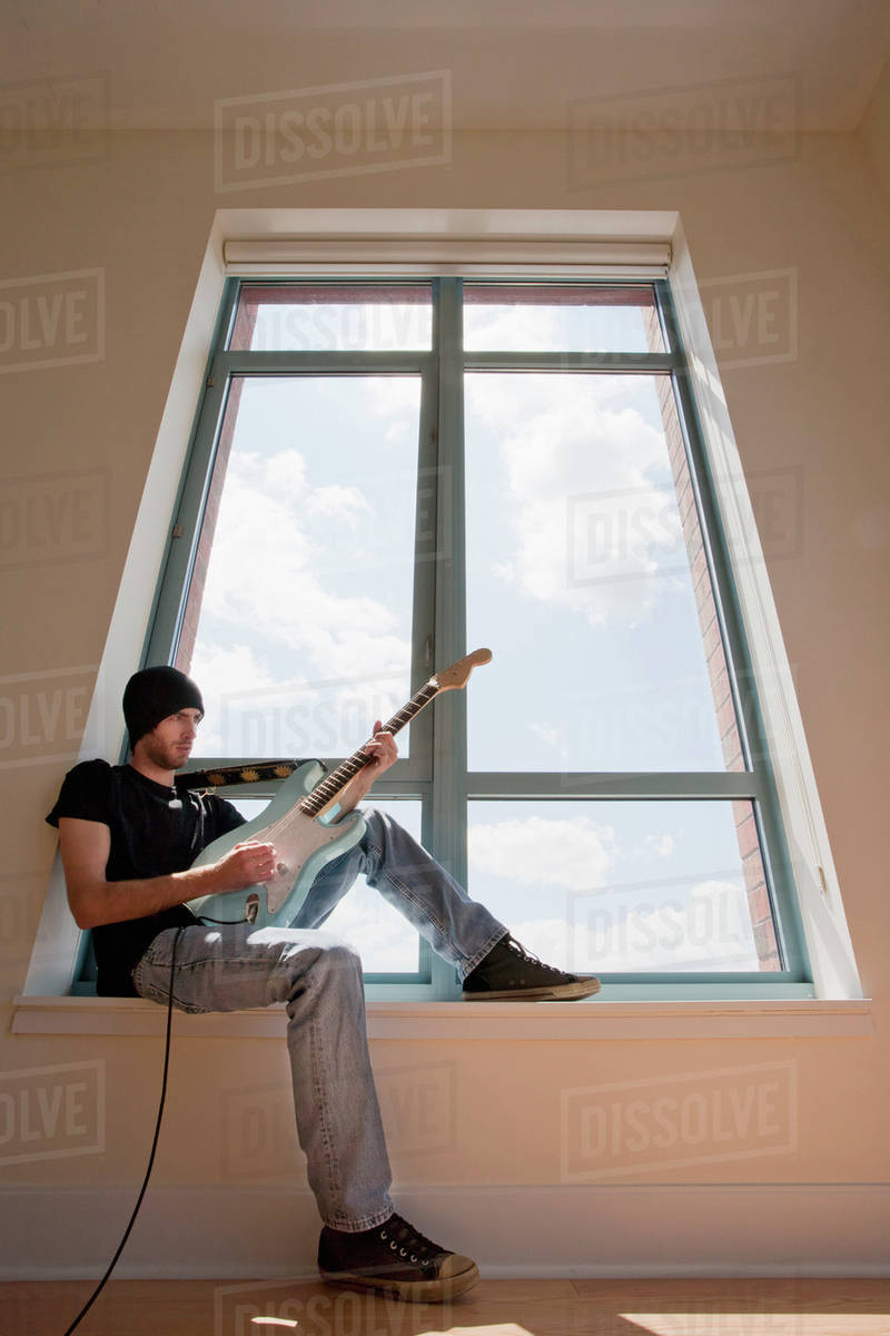 Low angle view of young man sitting on window sill and playing electric ...