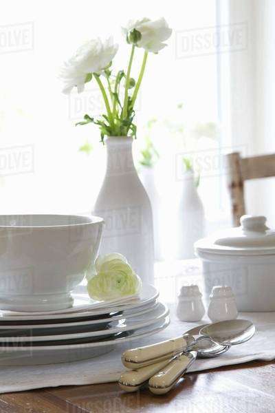 Stacked crockery and cutlery on a table with a vase of white ranunculus ...