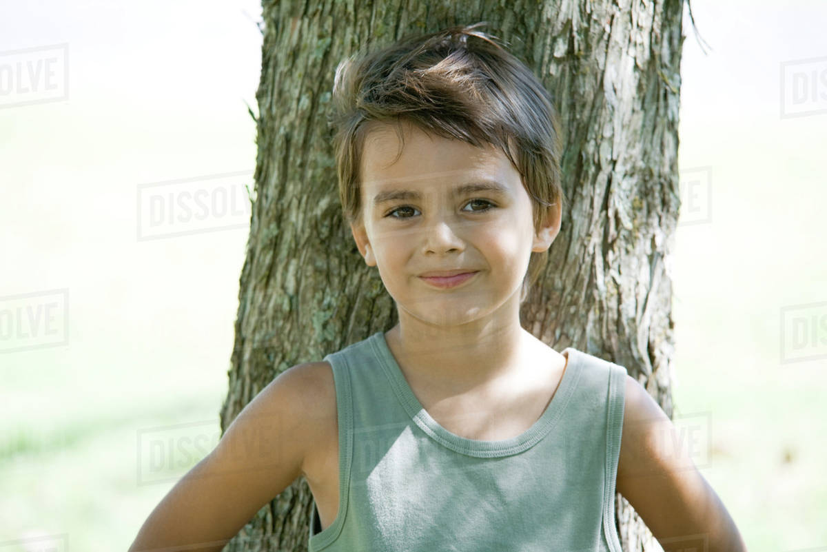 Little boy outdoors, leaning against tree trunk, smiling at camera ...