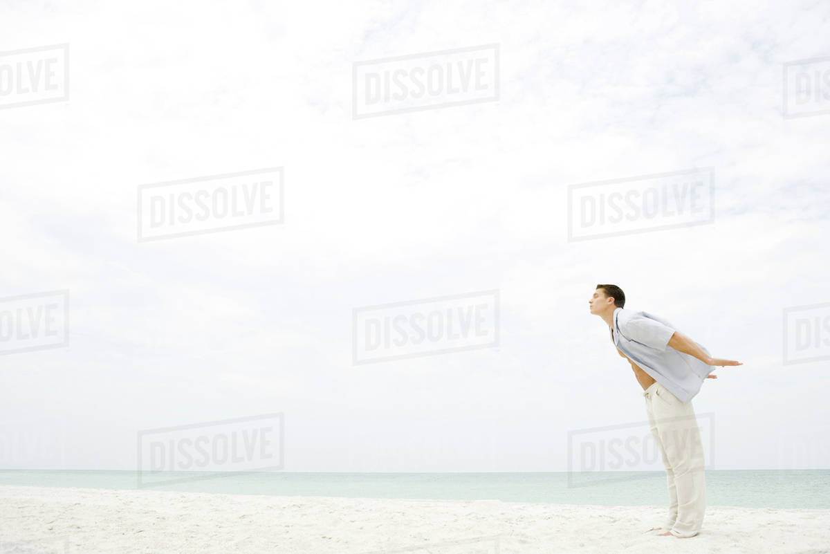 Man standing on beach, leaning into wind, arms behind back and eyes ...