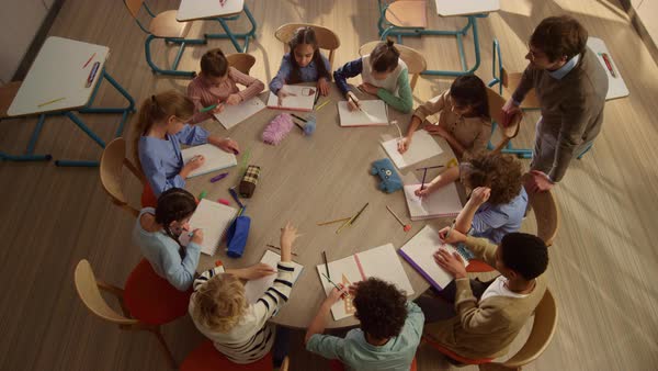 Schoolchildren having lesson at round table. Male teacher helping kids ...