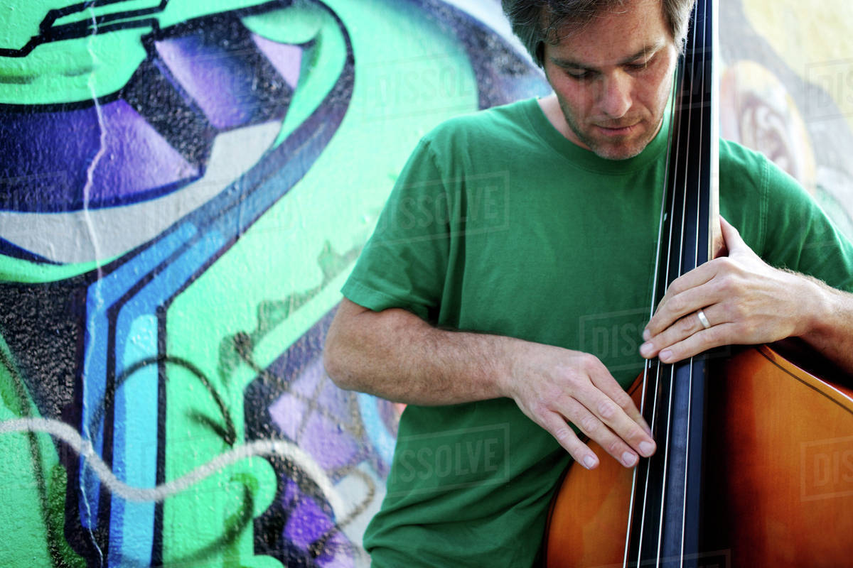Caucasian man playing double bass in front of graffiti covered wall ...