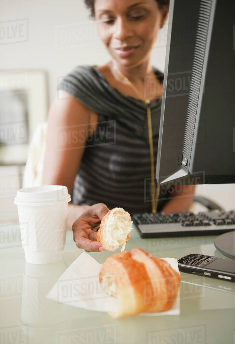 Black businesswoman eating croissant at desk - Royalty-free Stock Photo ...