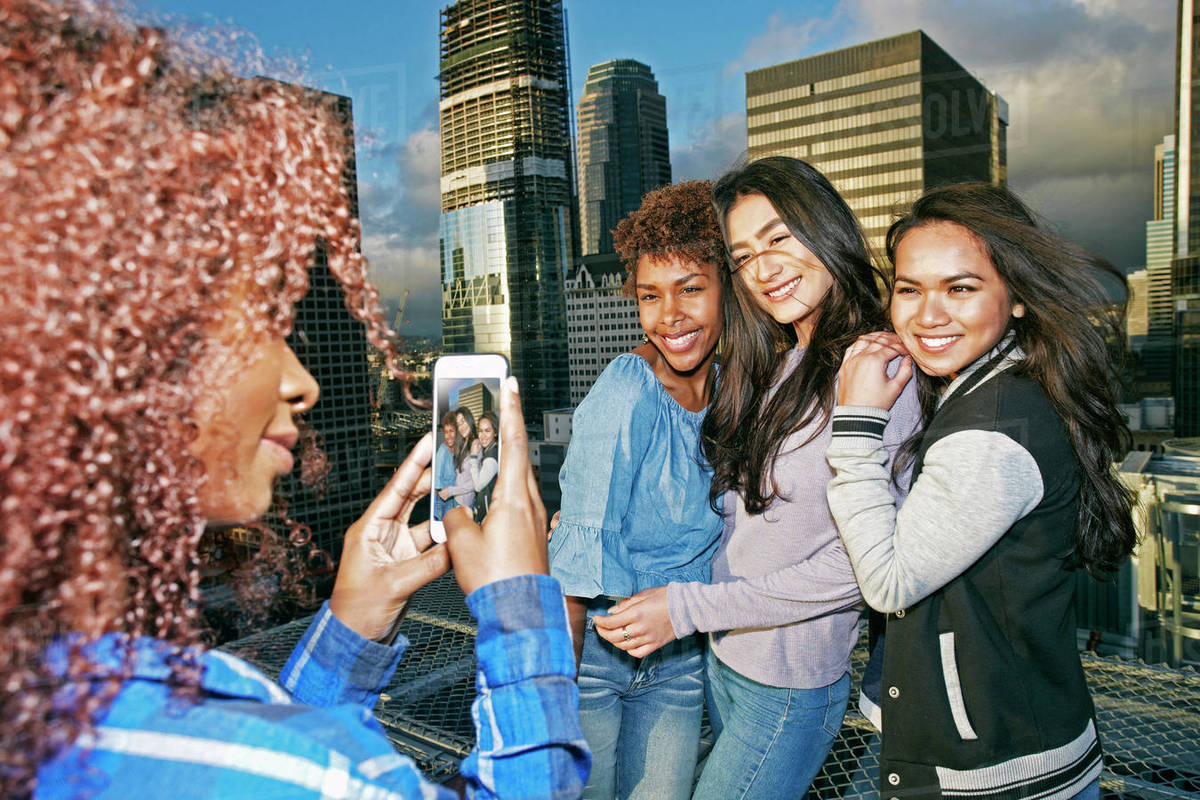 Stylish woman photographing friends on urban rooftop with cell phone ...