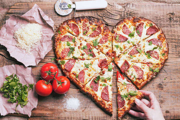 Hand pulling slice of heart-shaped pizza near ingredients on cutting ...