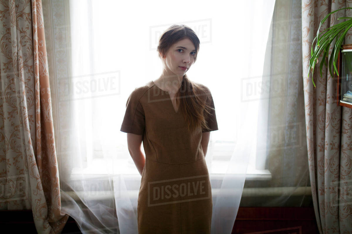 Serious Caucasian woman standing near window curtain - Royalty-free ...