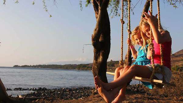 Two adorable little sisters sit on a rope swing together and wave at ...