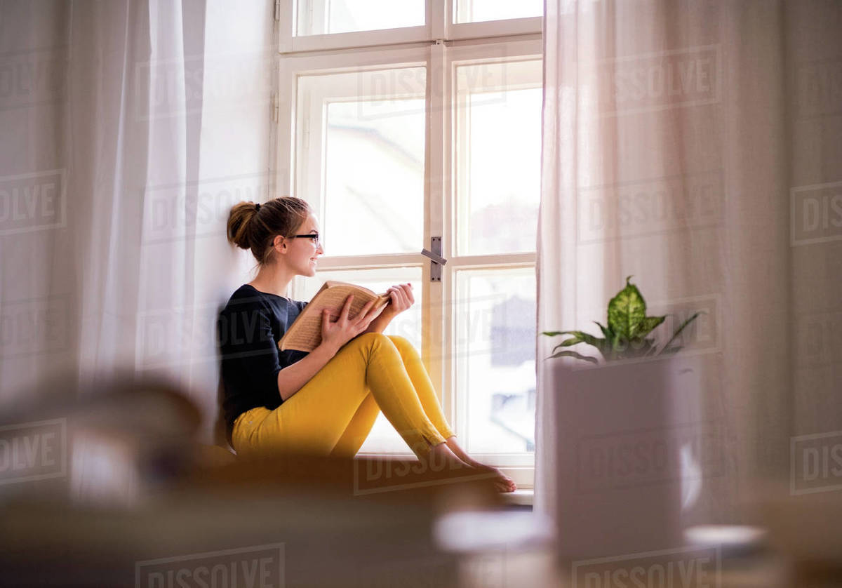 A young happy college female student with a book sitting on window sill ...
