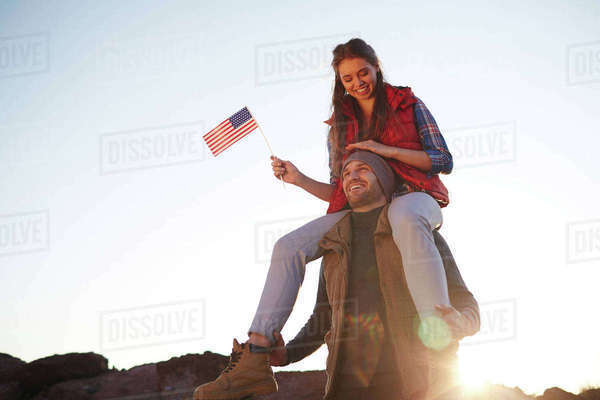 Happy young woman with American flag sitting on her boyfriend shoulders ...