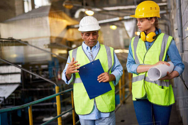 Two contemporary engineers in uniform and protective helmets discussing ...