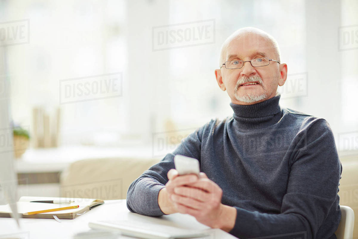 Older man with smartphone looking at camera by desk - Royalty-free ...
