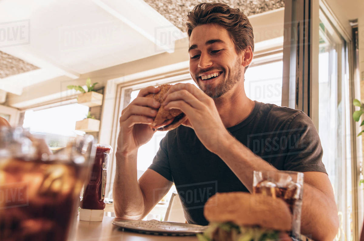 Happy young man eating a delicious burger at restaurant. Hungry young ...