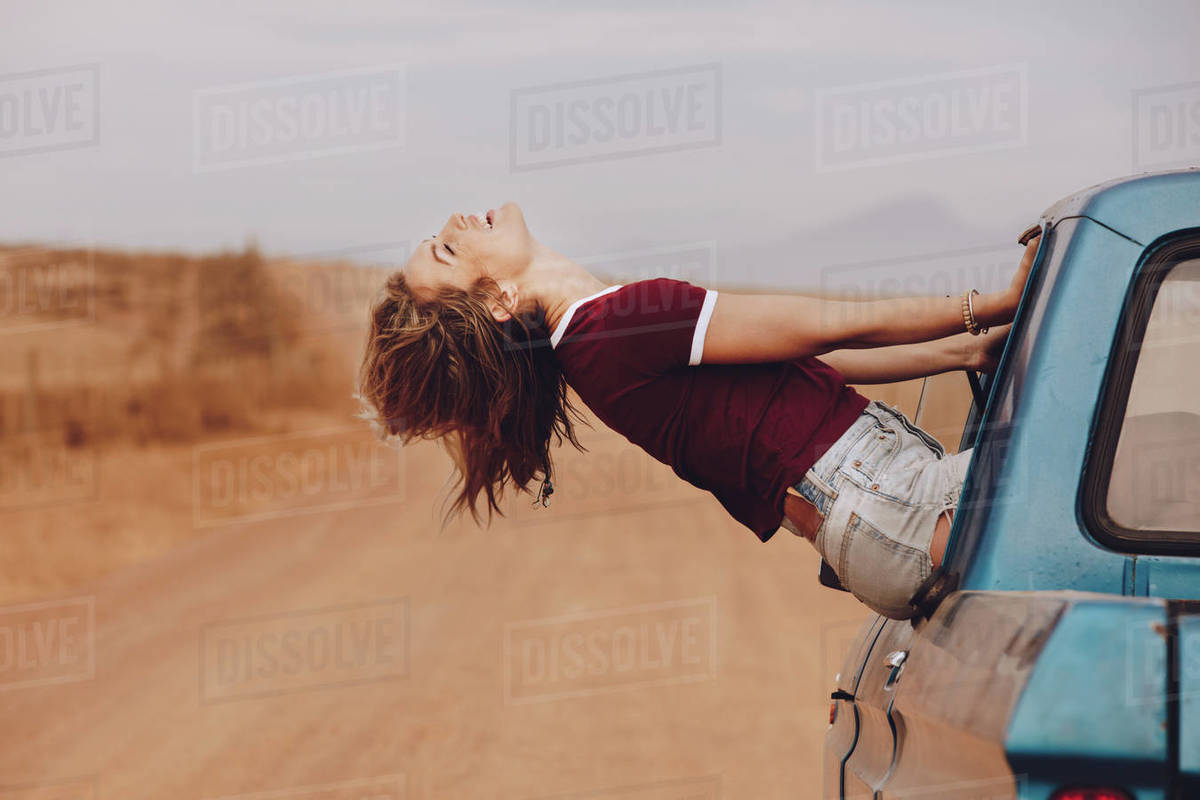 Beautiful young woman sitting on car window and looking up smiling ...