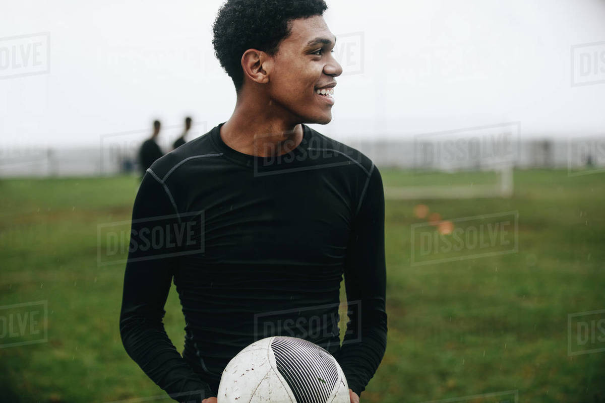 Smiling soccer player standing on field holding a football looking away ...