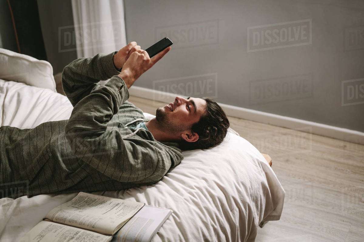 Student using mobile phone lying on bed with books by his side. Student ...