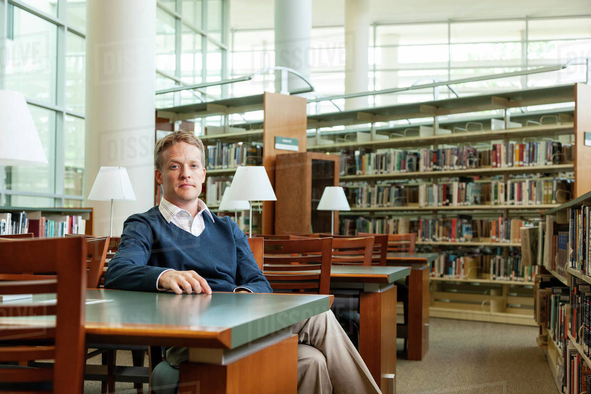 Portrait of college professor sitting at table in library looking at ...
