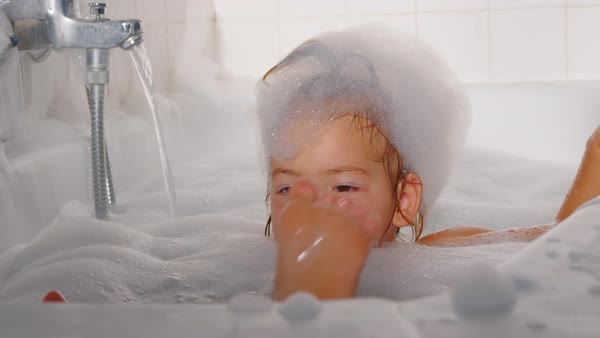 A little cheerful girl washes and plays in the bath with foam. Daily ...