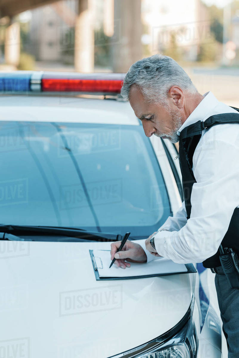 side view of focused male police officer writing in clipboard on car at ...