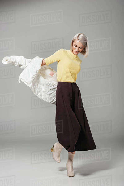 smiling young woman putting on stylish white jacket on grey background ...