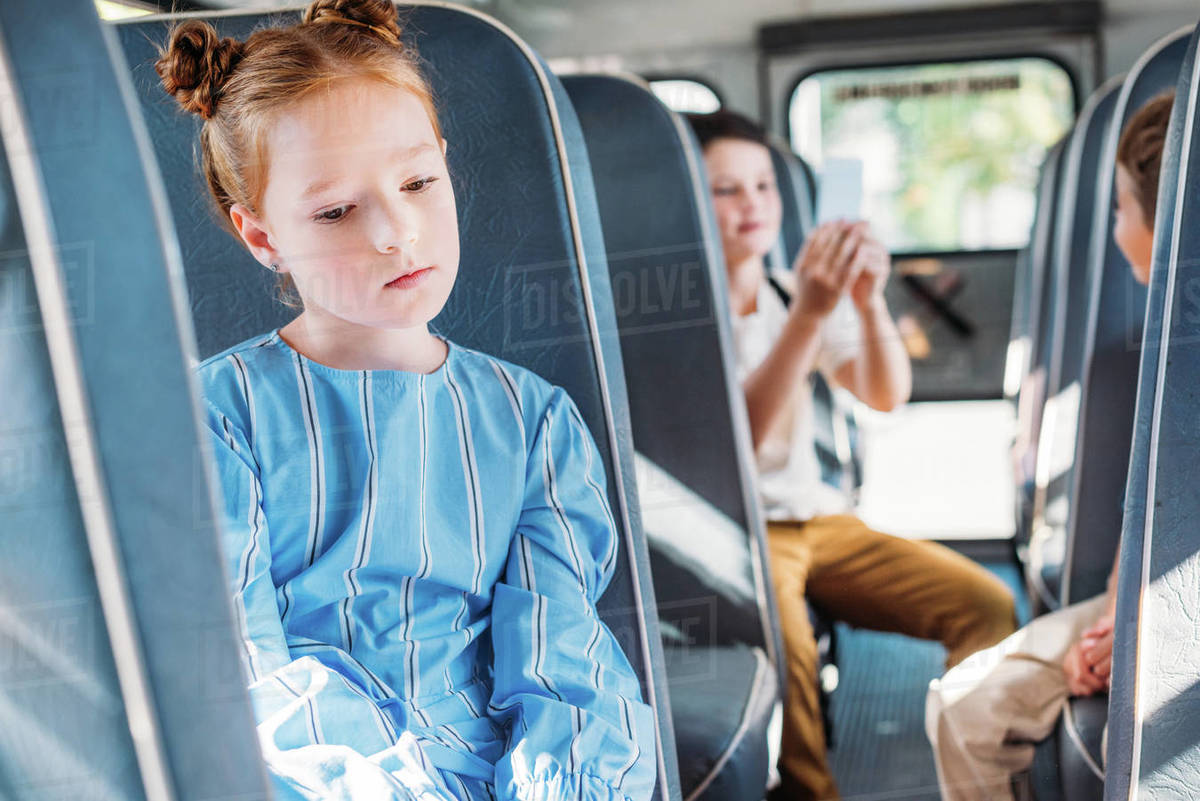 sad little schoolgirl sitting alone in school bus while her classmates ...