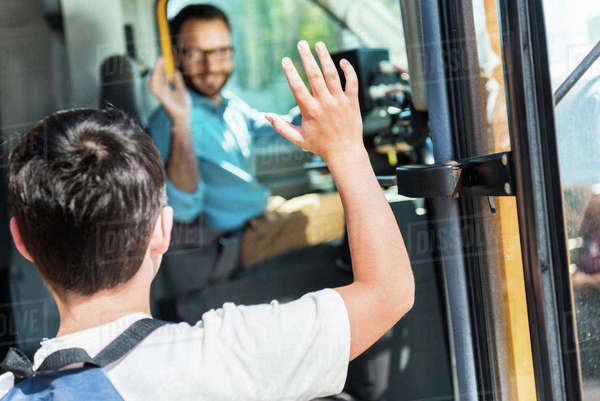 rear view of schoolboy waving to happy bus driver while entering bus ...