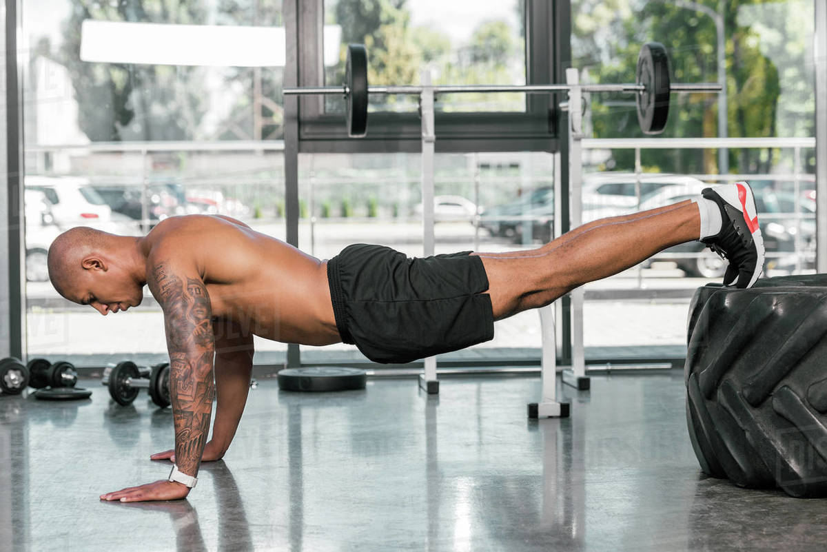 side view of muscular african american man doing push ups with tyre in ...