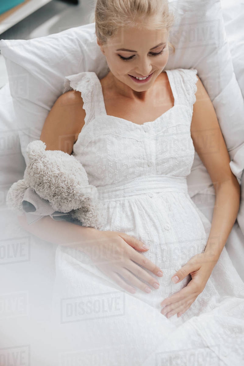 Overhead view of smiling pregnant woman in white nightie with teddy ...