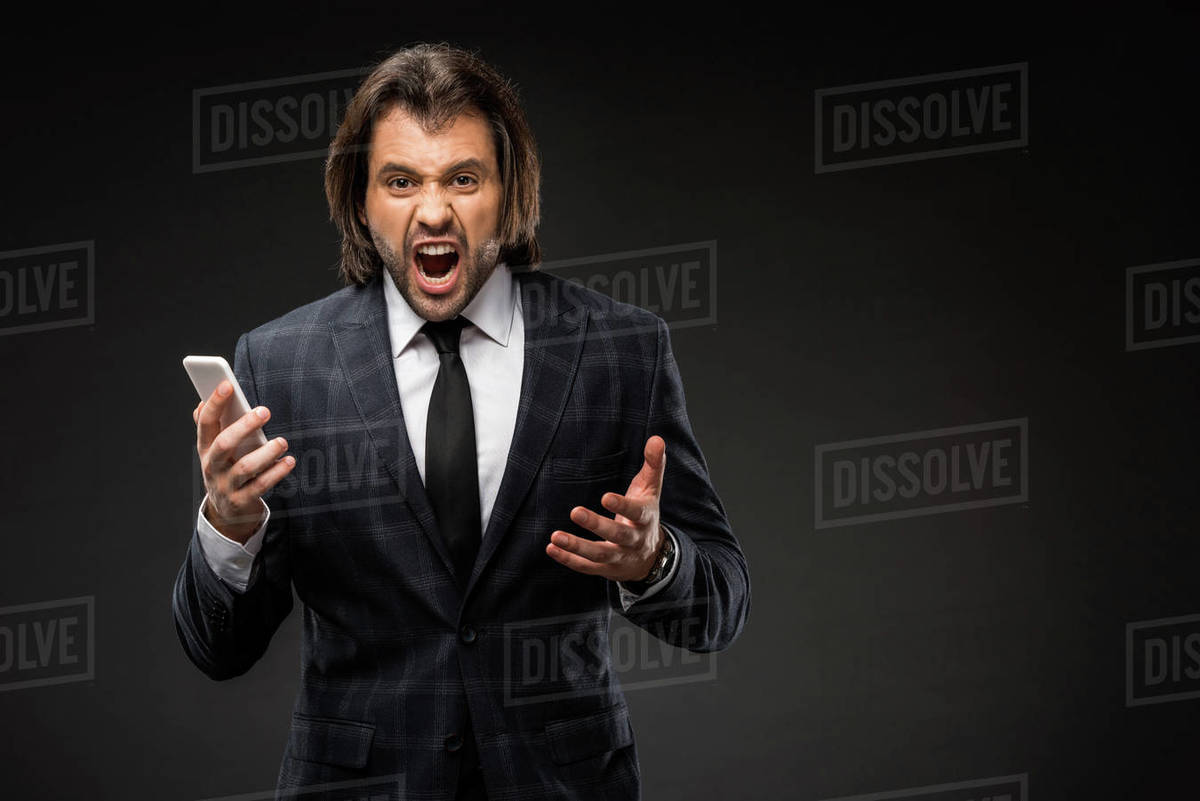 Angry young businessman holding smartphone and yelling at camera ...