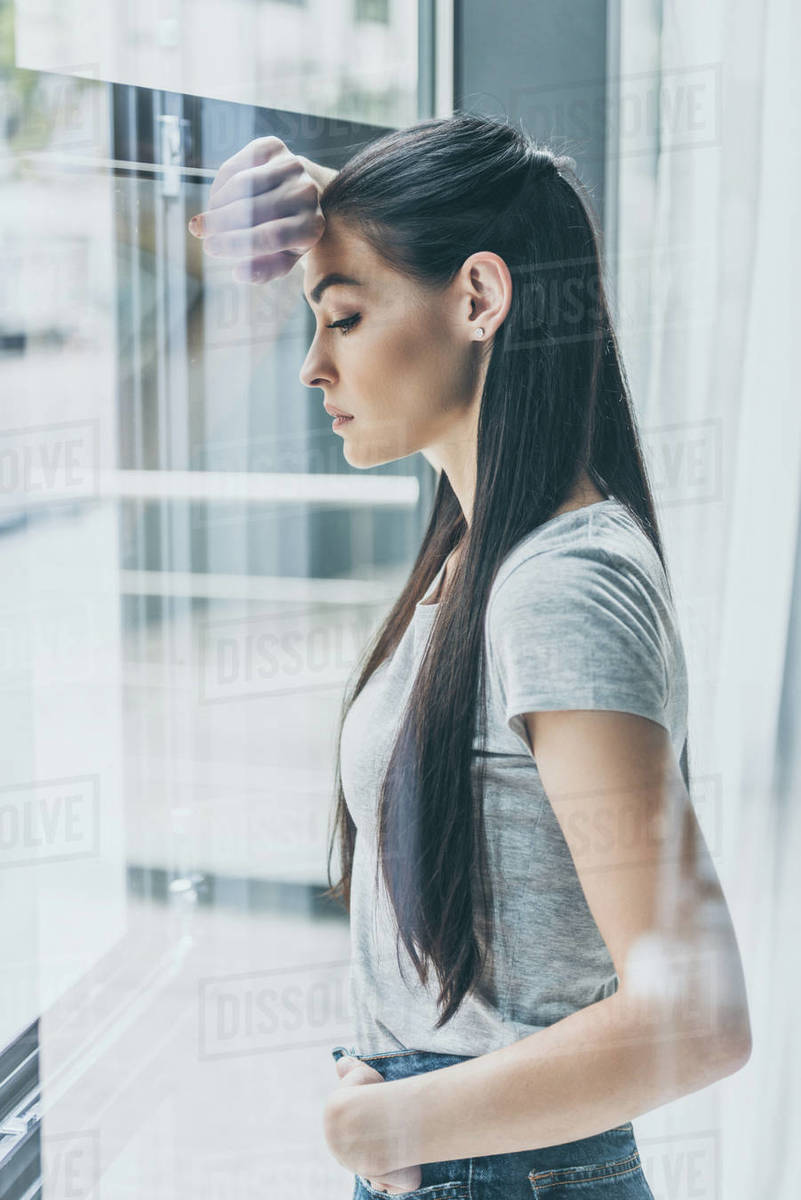 Side view of upset young woman leaning at window and looking down ...