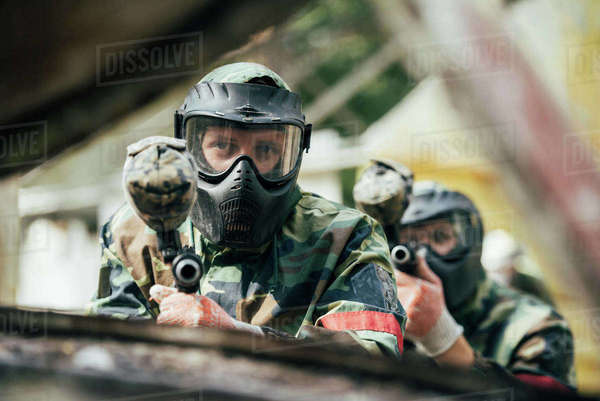 Male paintballer and his team in uniform and protective masks aiming by ...