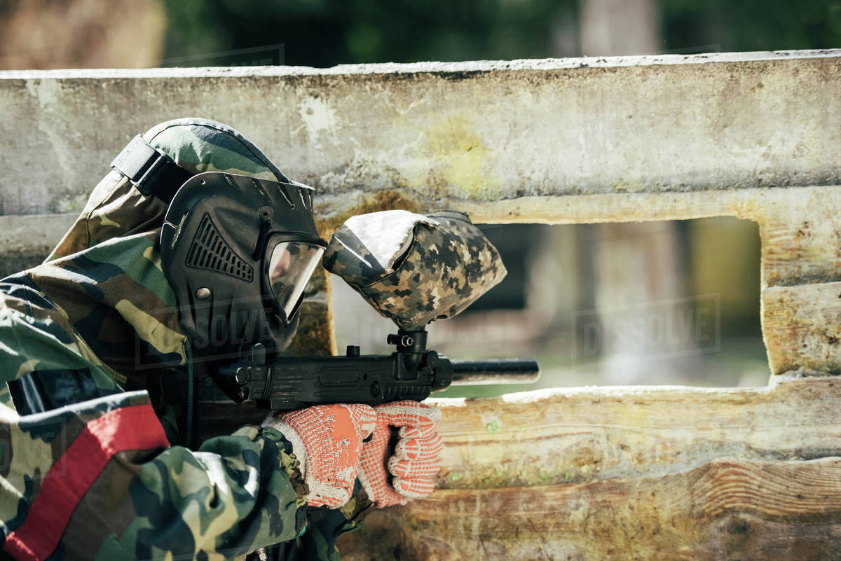 Rear view of paintball player in goggle mask and camouflage aiming by ...