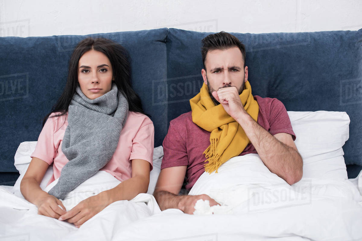 Sick young couple with cough sitting in bed and looking at camera ...
