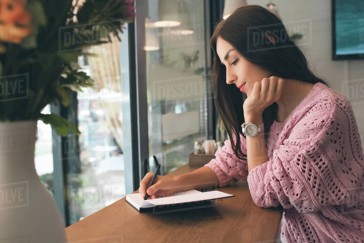 Side view of young woman making notes in textbook at table in cafe ...