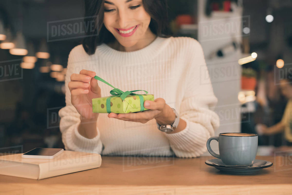 Smiling young woman untying gift box at table with smartphone and coffee cup in cafe - Stock ...