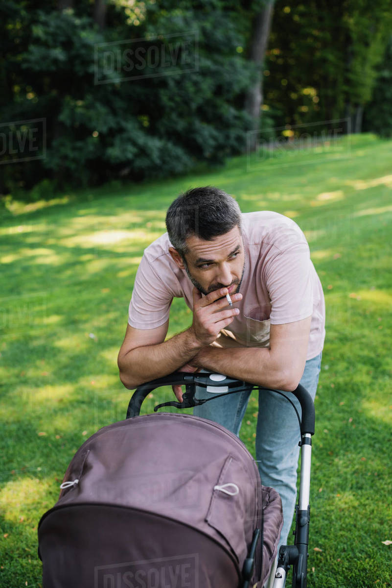Father smoking cigarette near baby carriage in park and looking away ...