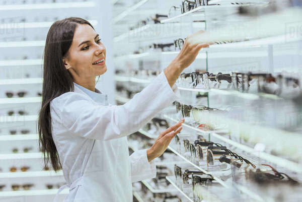 Professional smiling optician taking glasses from shelves in ...