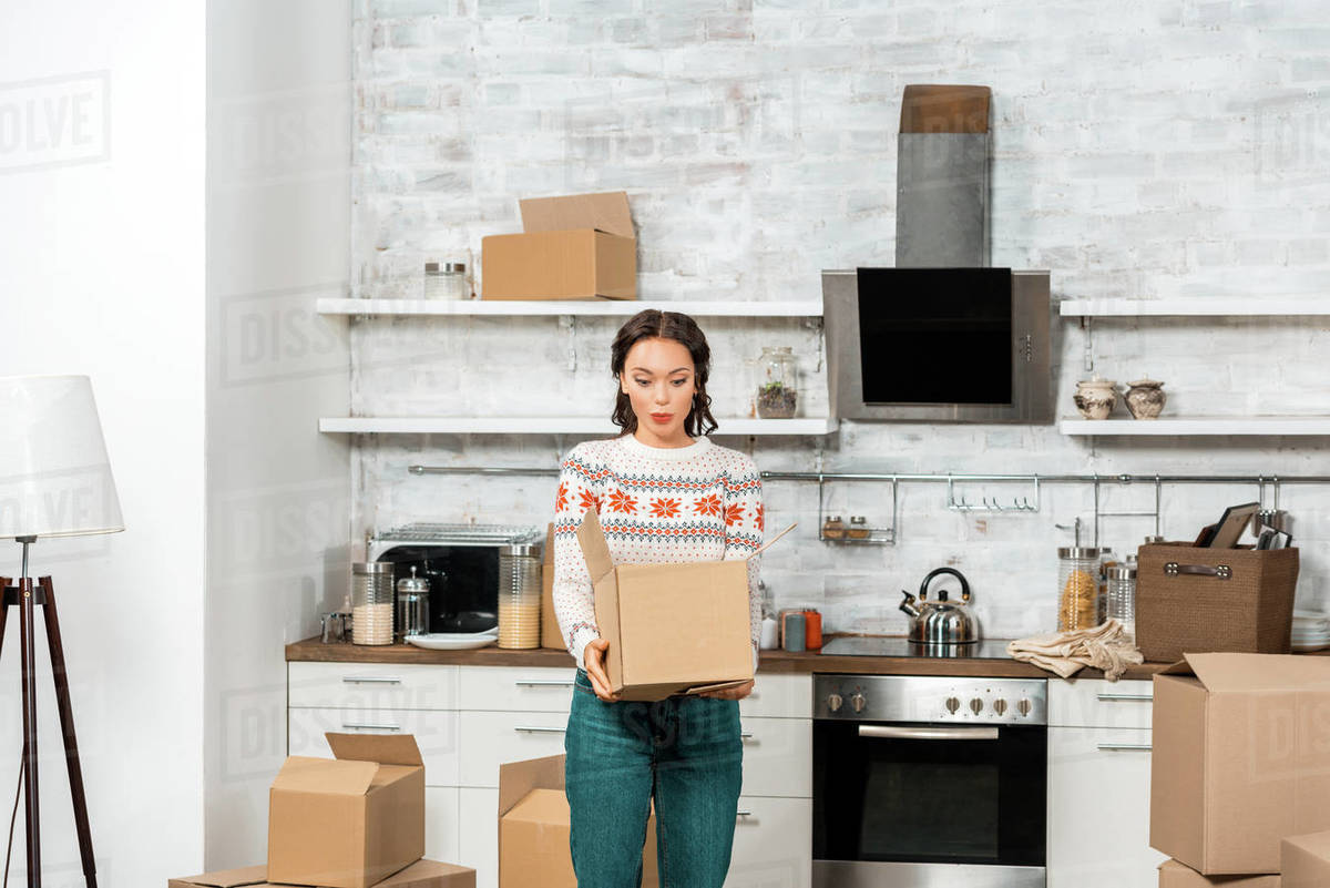 surprised young woman looking into cardboard box in kitchen at new home ...
