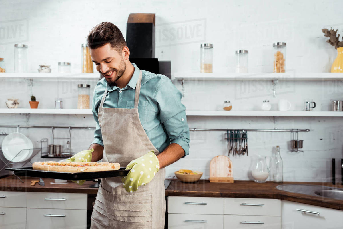 Smiling young man in apron and potholders holding baking tray with ...