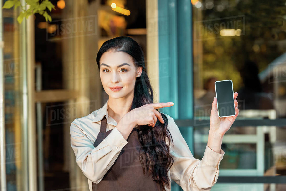 Beautiful waitress in apron pointing on smartphone with blank screen ...