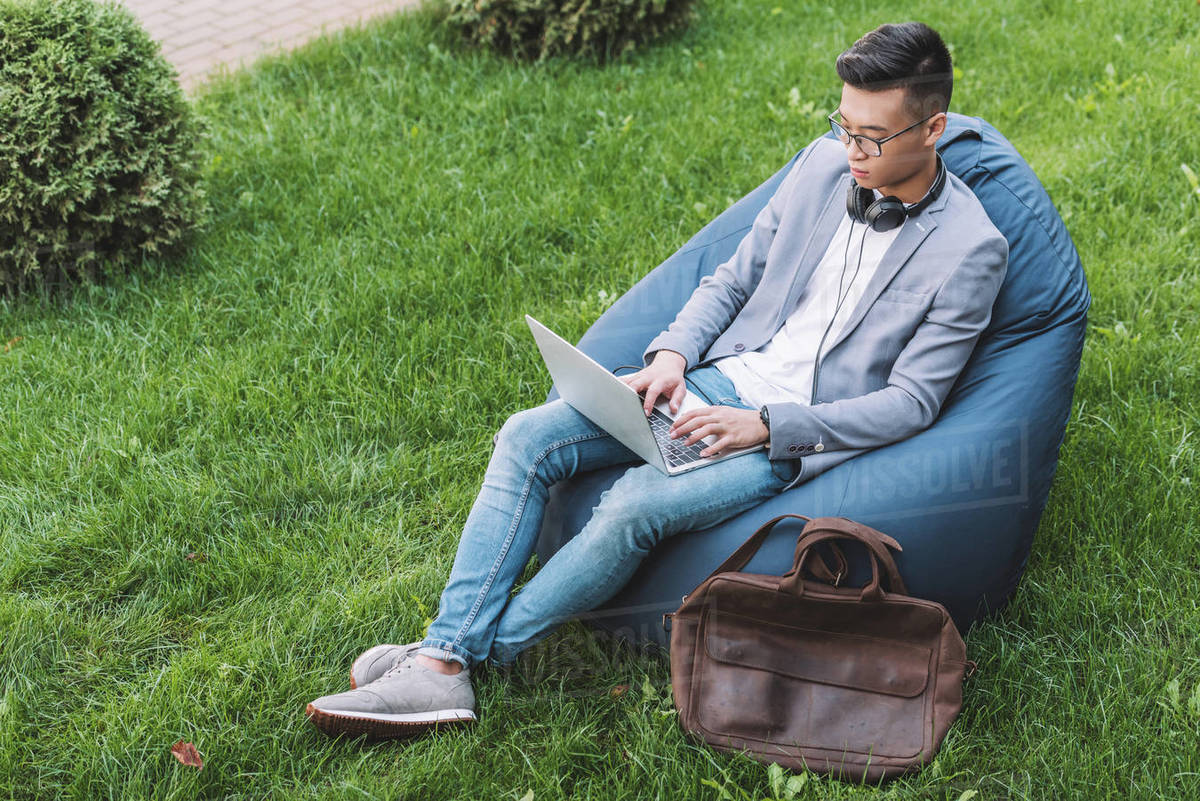 Handsome Asian teleworker working on laptop while sitting on bean bag ...