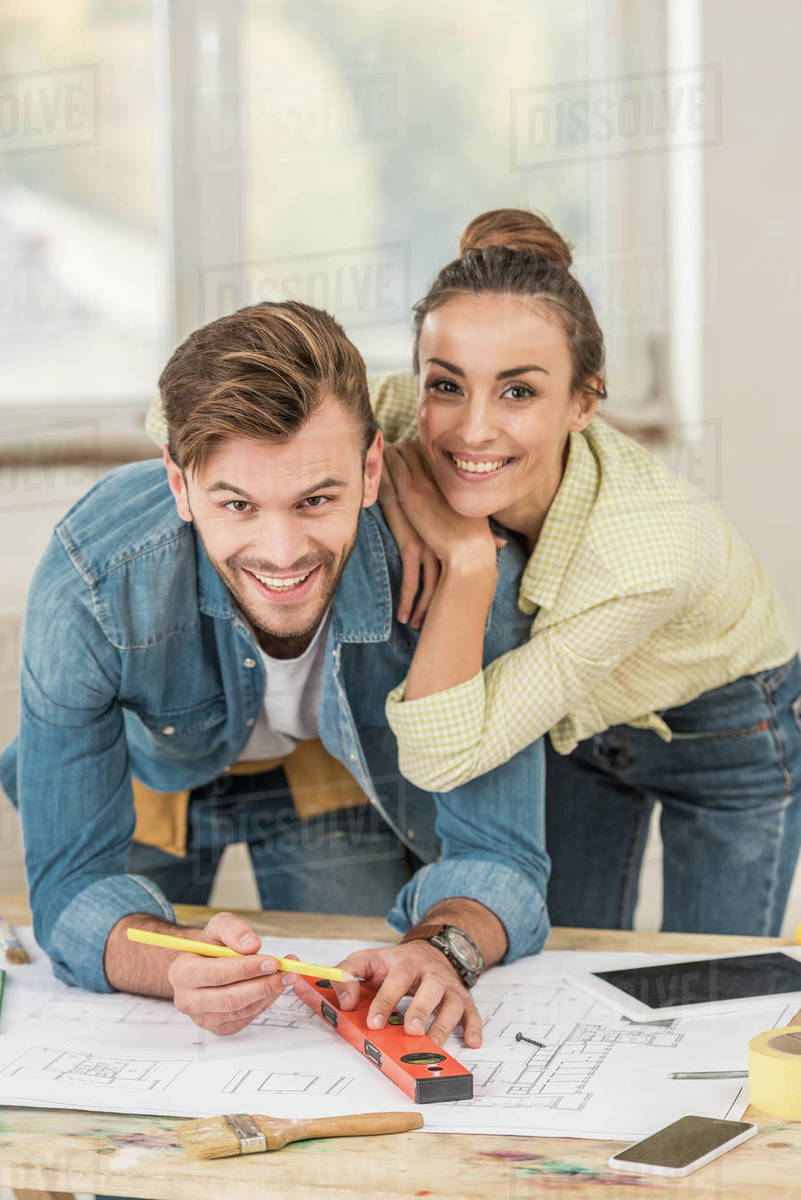 Happy young couple working with blueprint and smiling at camera during ...