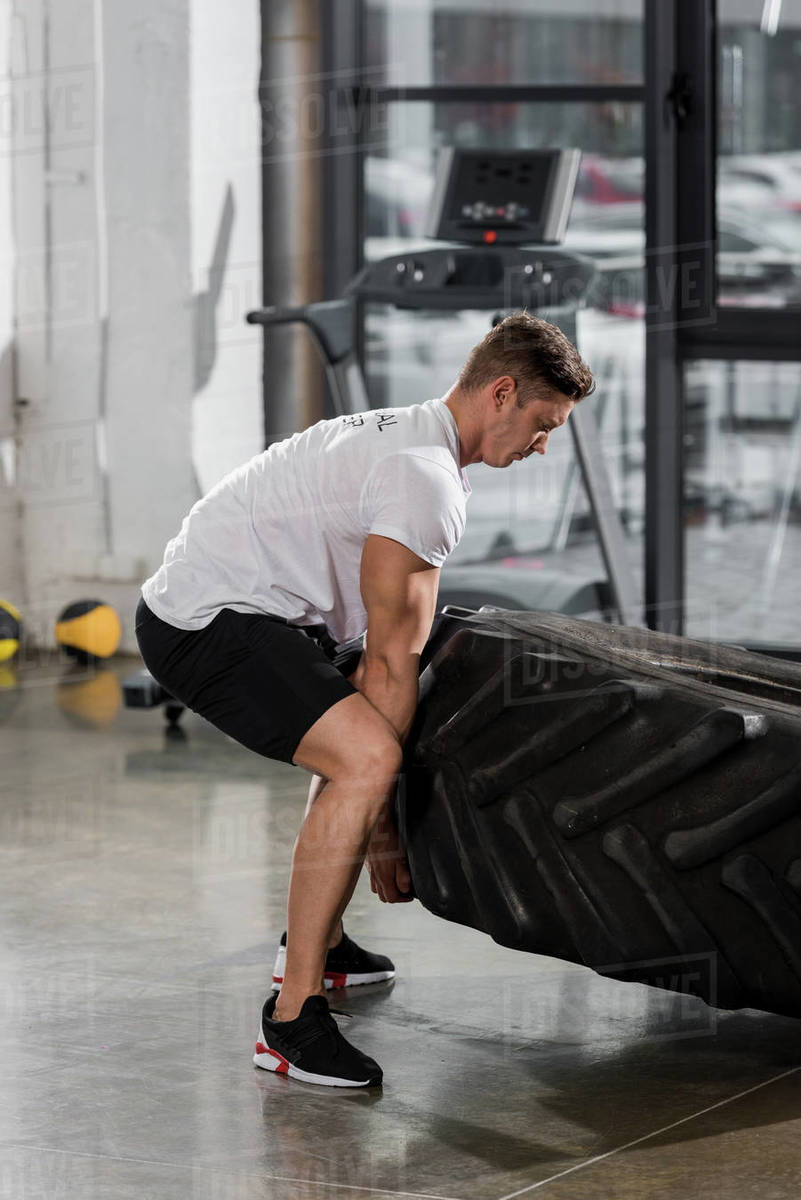 Side view of handsome muscular bodybuilder exercising with tire in gym ...