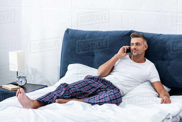 Cheerful adult man talking on smartphone while laying in bed at home ...